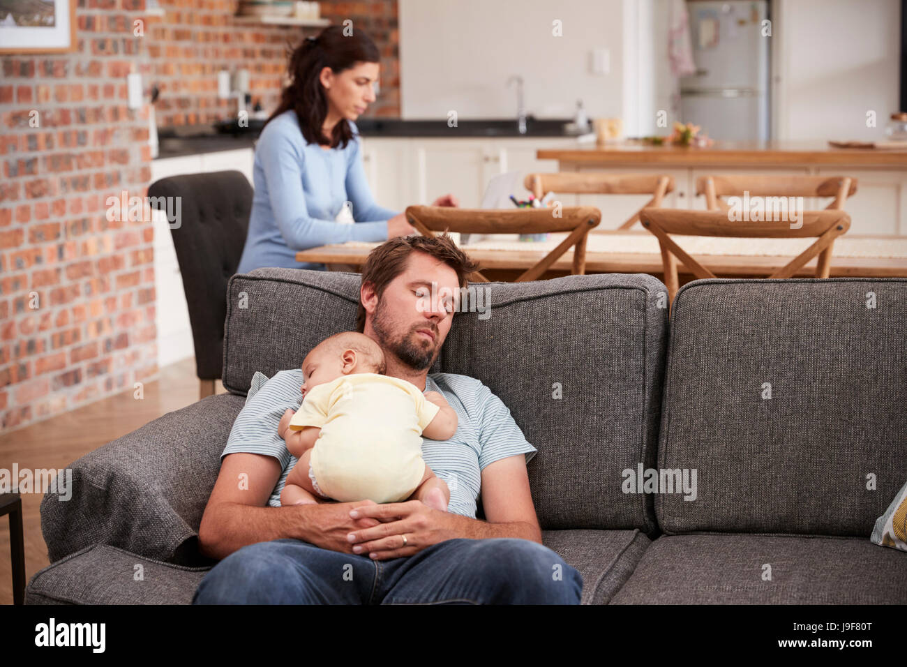 Tired Father With Baby Son Asleep On Sofa As Mother Works Stock Photo ...