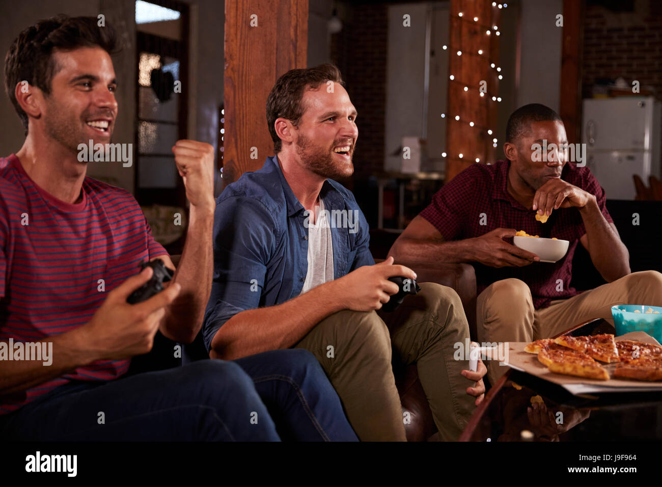 Three male friends playing video games and eating at home Stock Photo ...