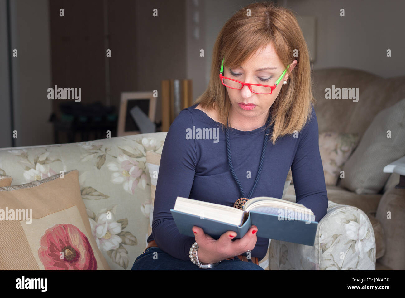 A mid age woman reading a book sitting at the living room of her home ...