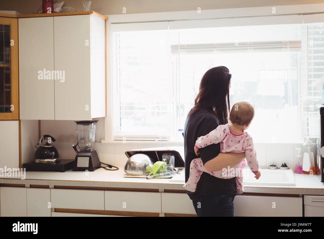 Rear view of mother and baby looking through kitchen window at home ...