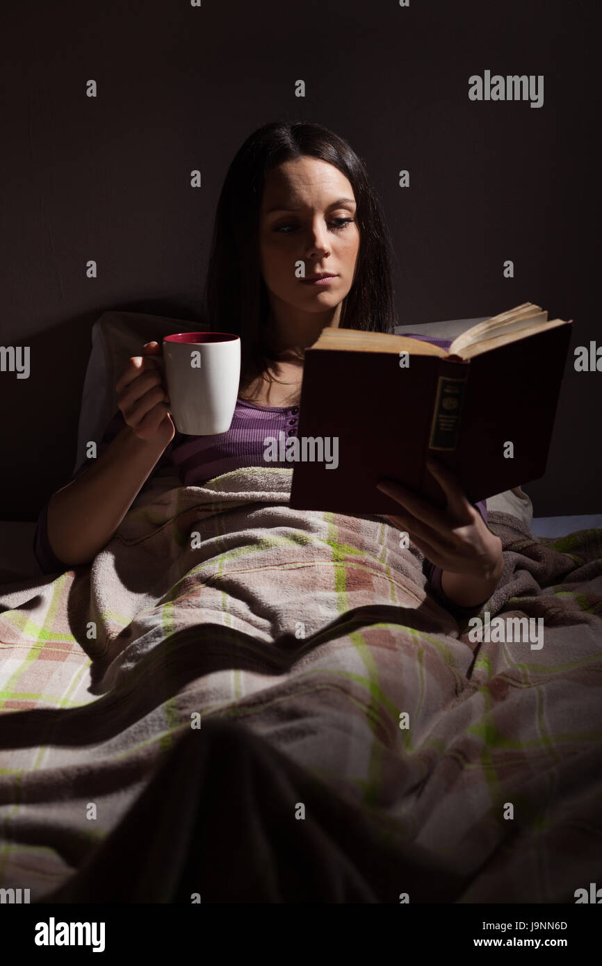 Young woman relaxing in her bed before sleep. She is reading a book and ...