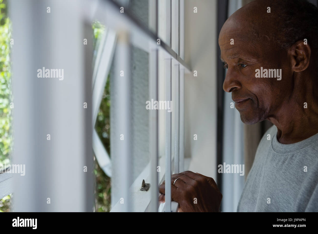 Thoughtful senior man looking out through window while standing at home ...