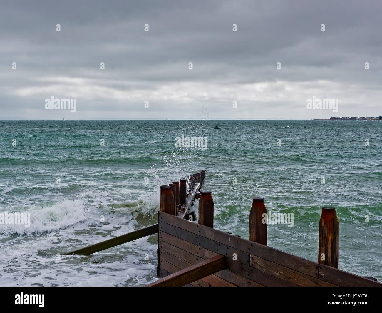 On a windy day, under a grey sky, wave crash against a partially ...