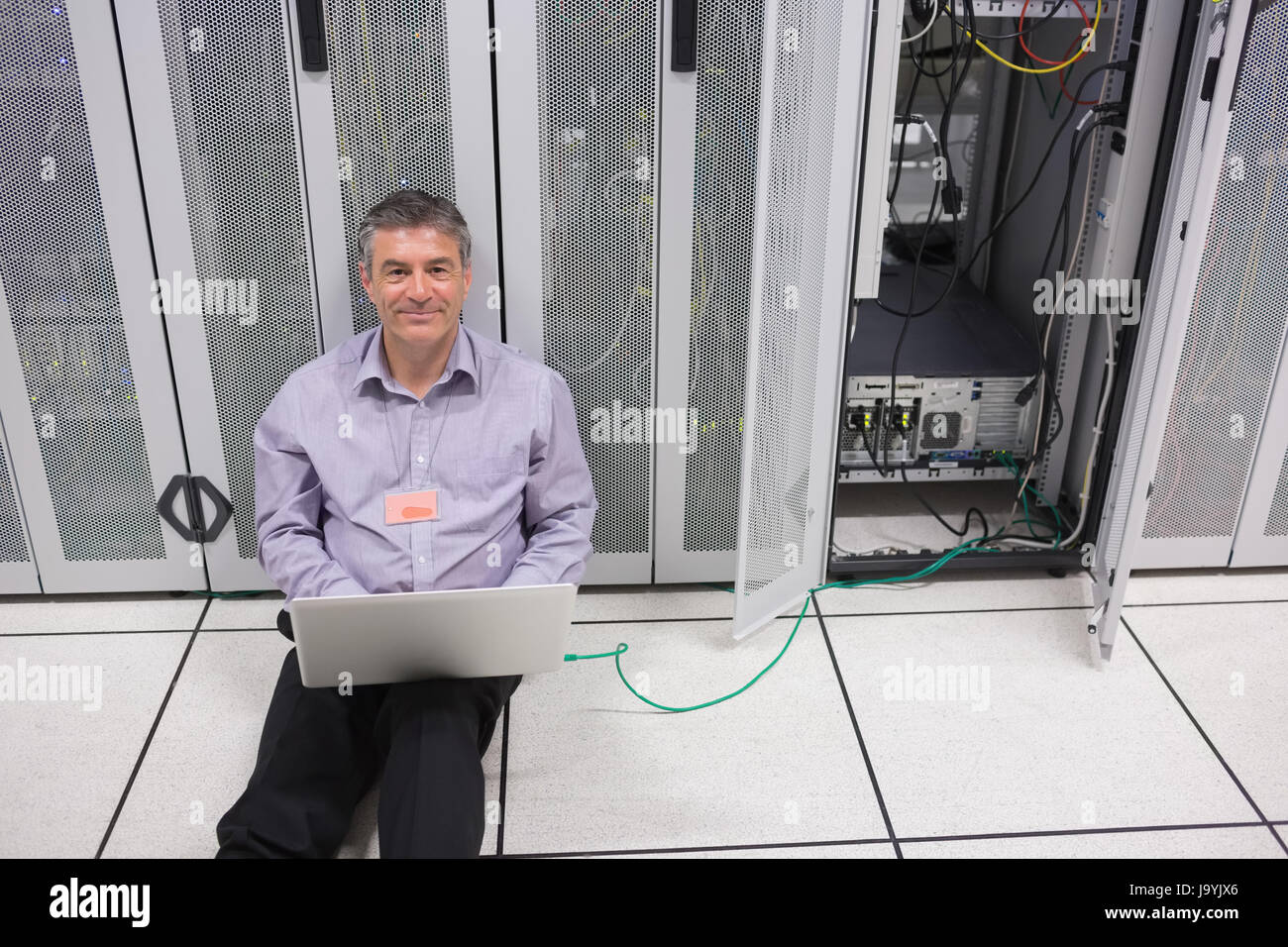 Smiling man doing server maintenance with laptop sitting on floor of ...