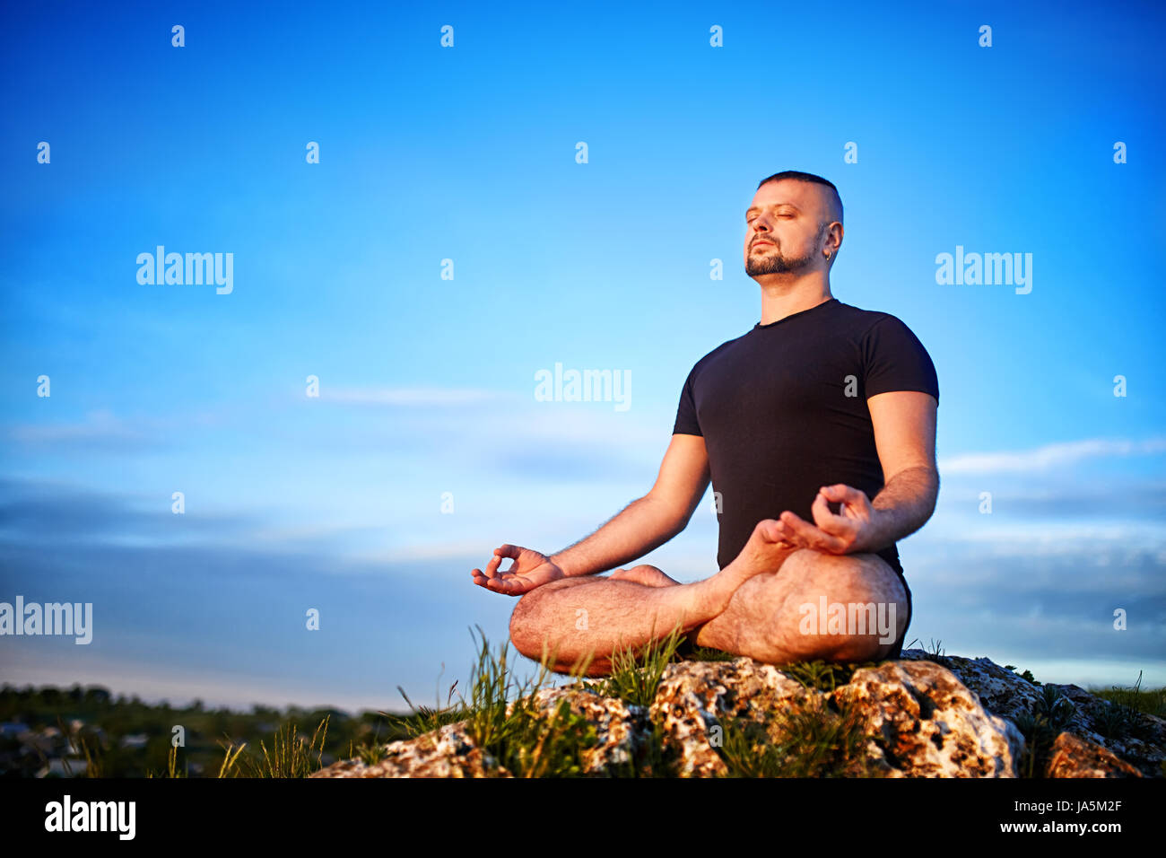 Attractive young man doing yoga on the rock against blue sky with ...