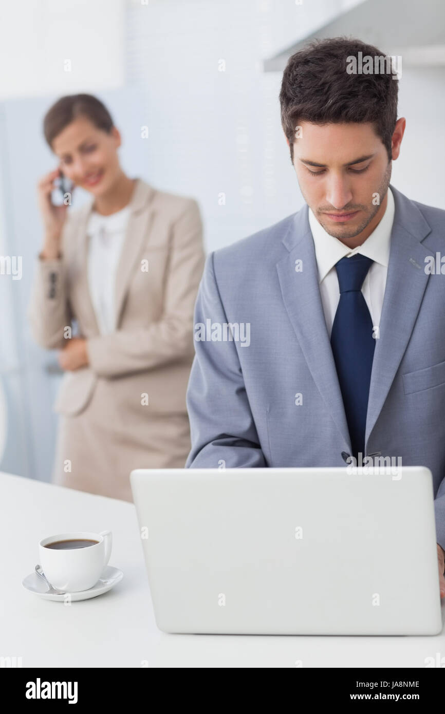 Businessman Using A Laptop Before Going To Work While His Wife Is On The Phone On The Background