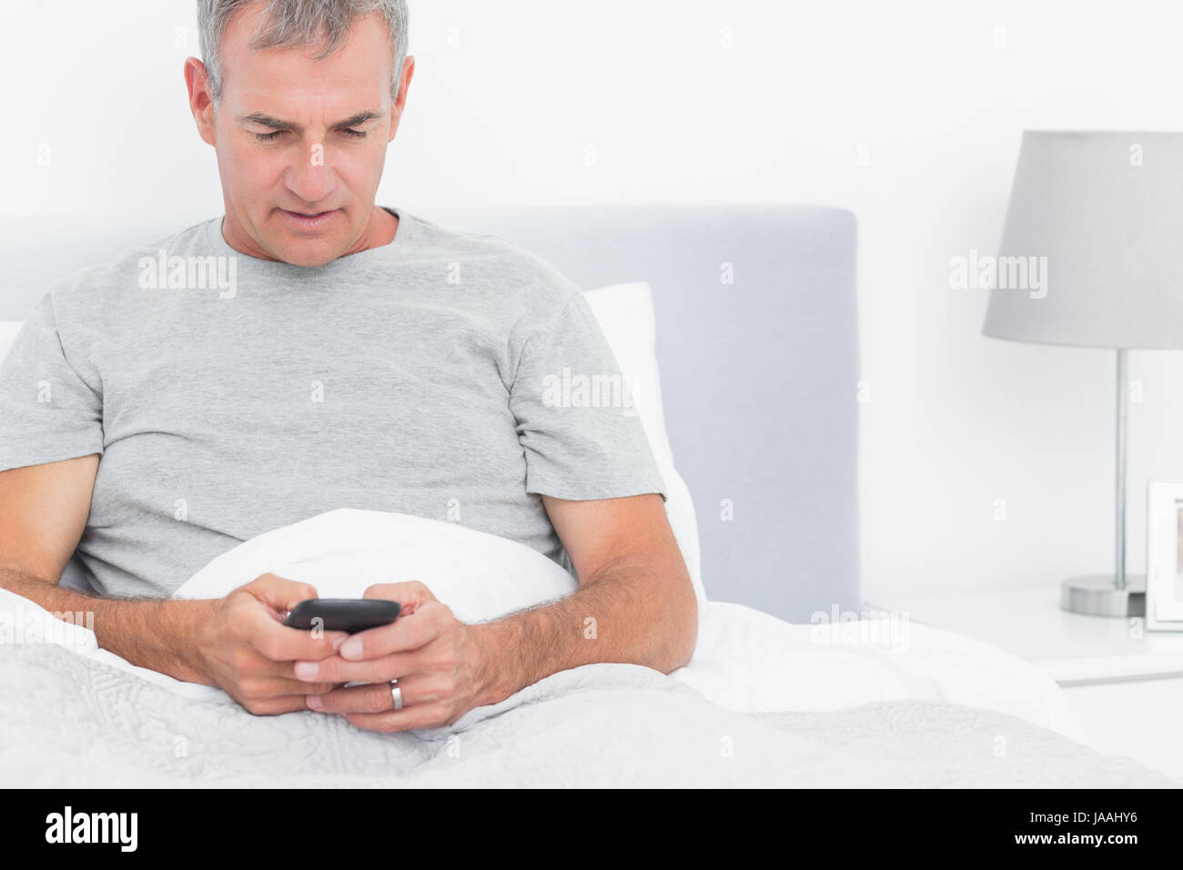 Grey haired man sending a text in bed in bedroom at home Stock Photo ...