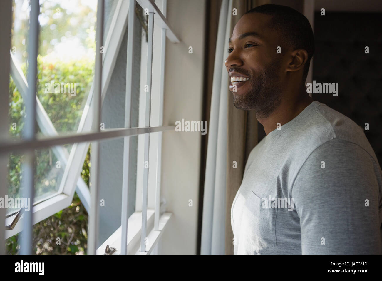 Thoughtful smiling man looking out through window while standing at ...