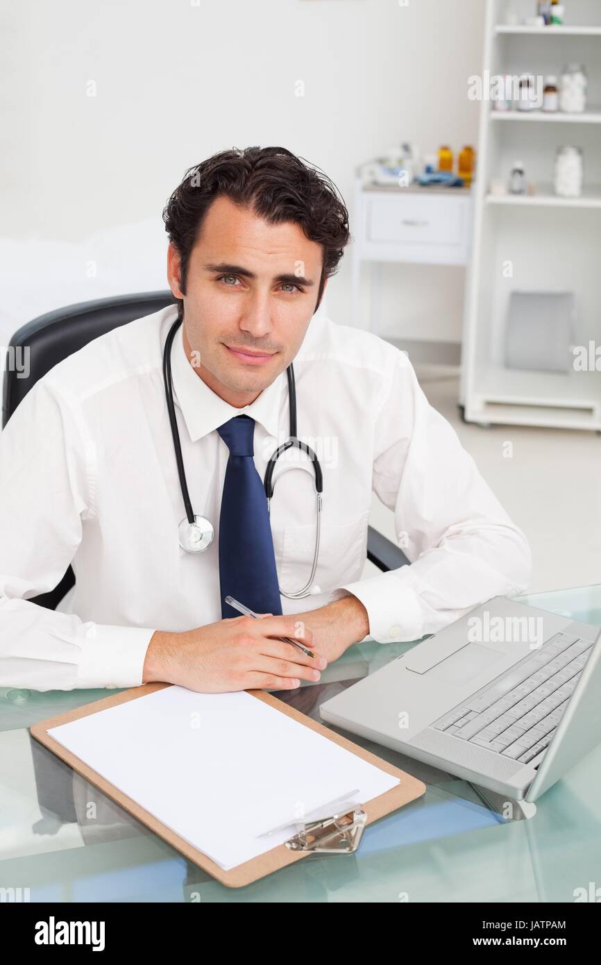 Doctor sitting at his desk with clipboard and laptop Stock Photo - Alamy