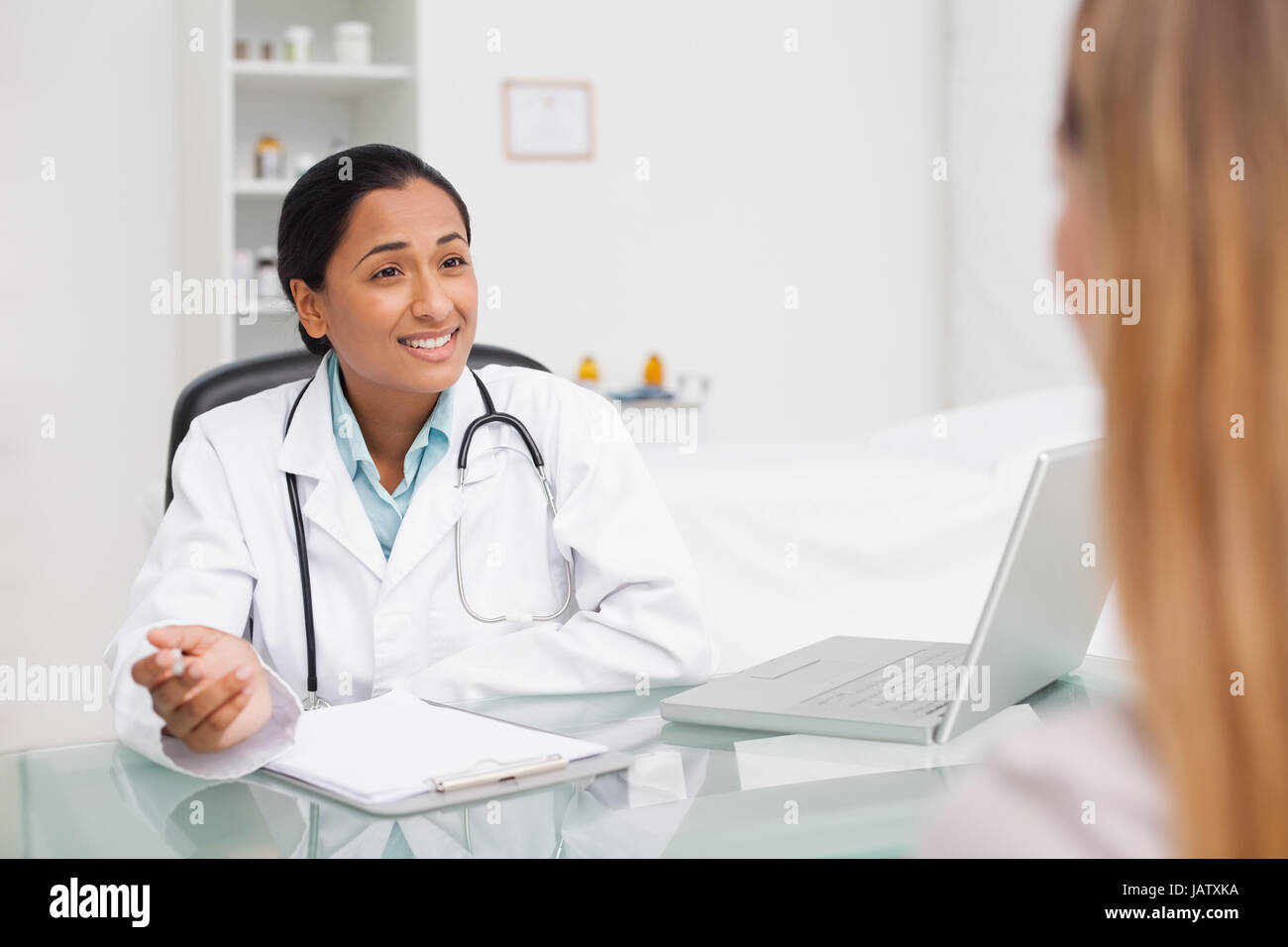 Practitioner sitting a desk with a clipboard and a laptop while talking ...