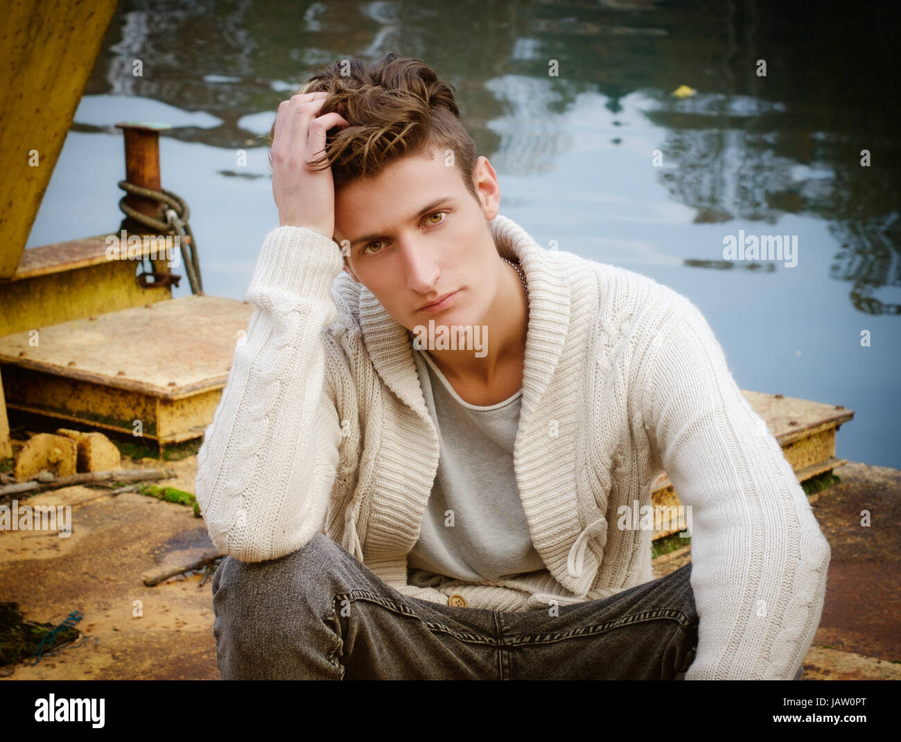 Attractive young man sitting on rusty metal next to river waters ...