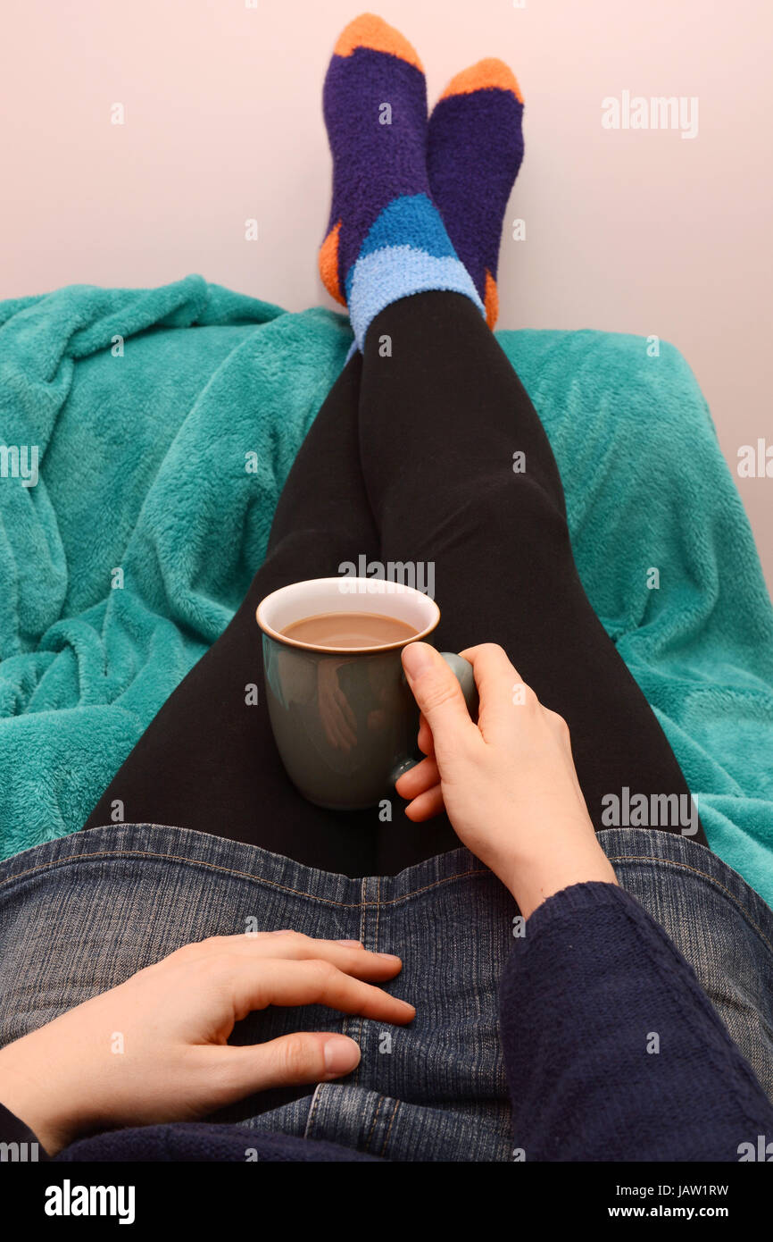 Woman dressed warmly, holding hot coffee or tea, with her feet up on ...