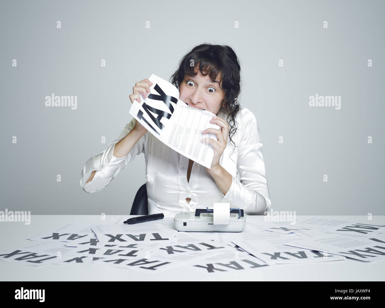 Young desperate woman at her paperwork covered desk biting a tax form ...