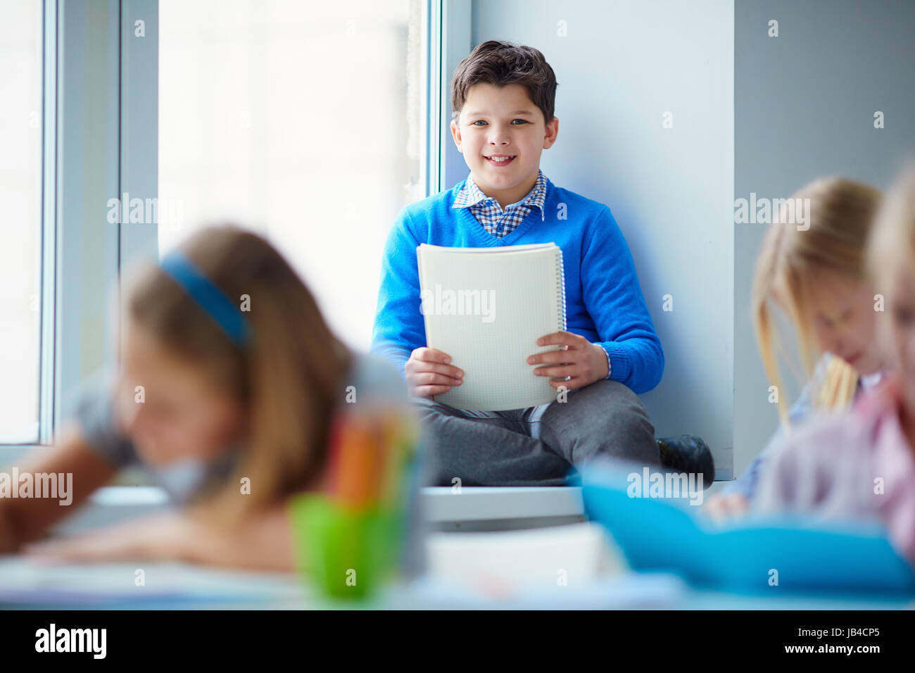 Portrait of happy schoolboy sitting by the classroom window and looking ...