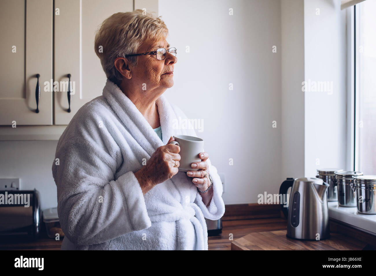 Senior woman looking out of her kitchen window with a cup of tea in ...