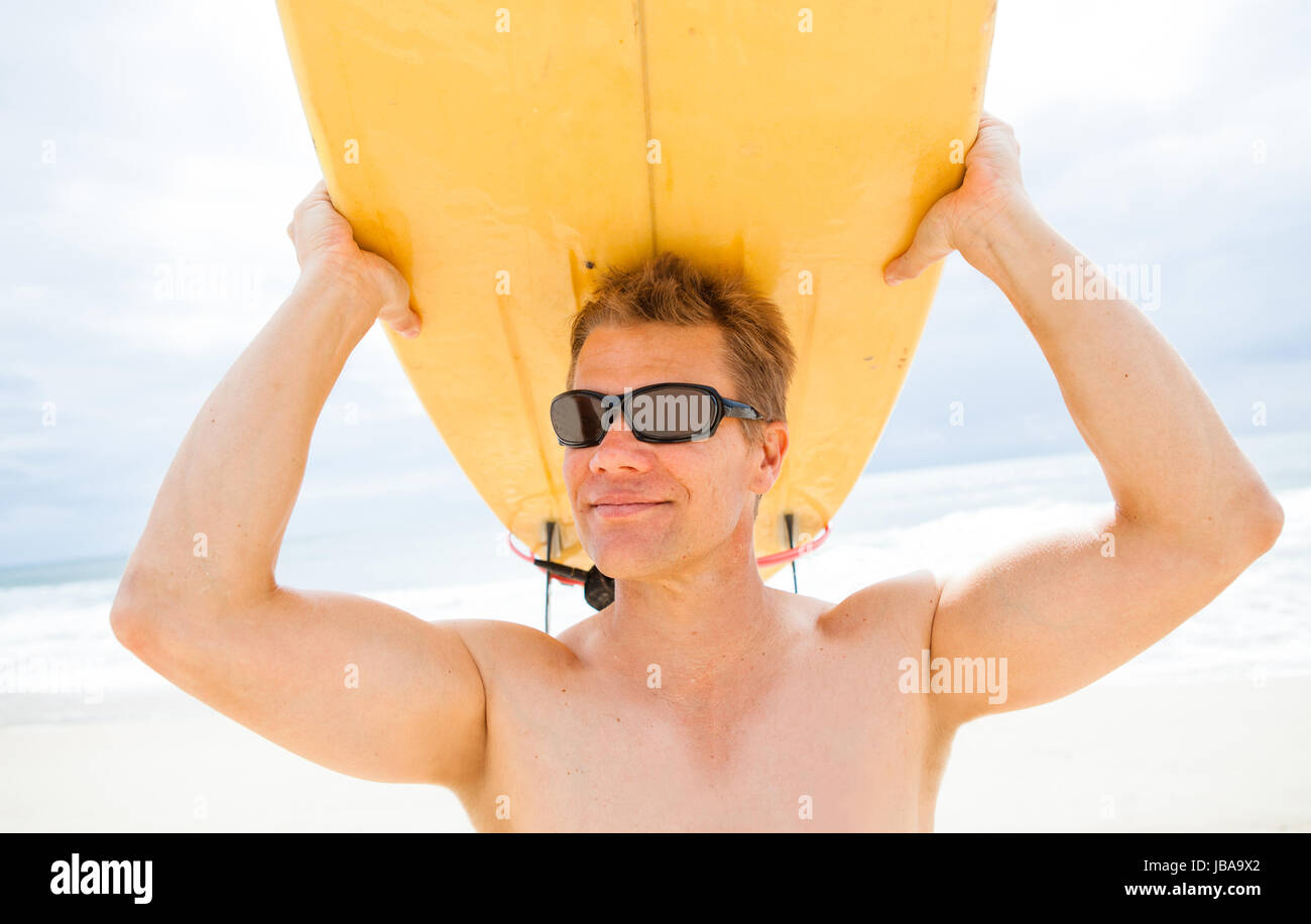 Smiling male surfer resting surfboard on head at beach with ocean and ...