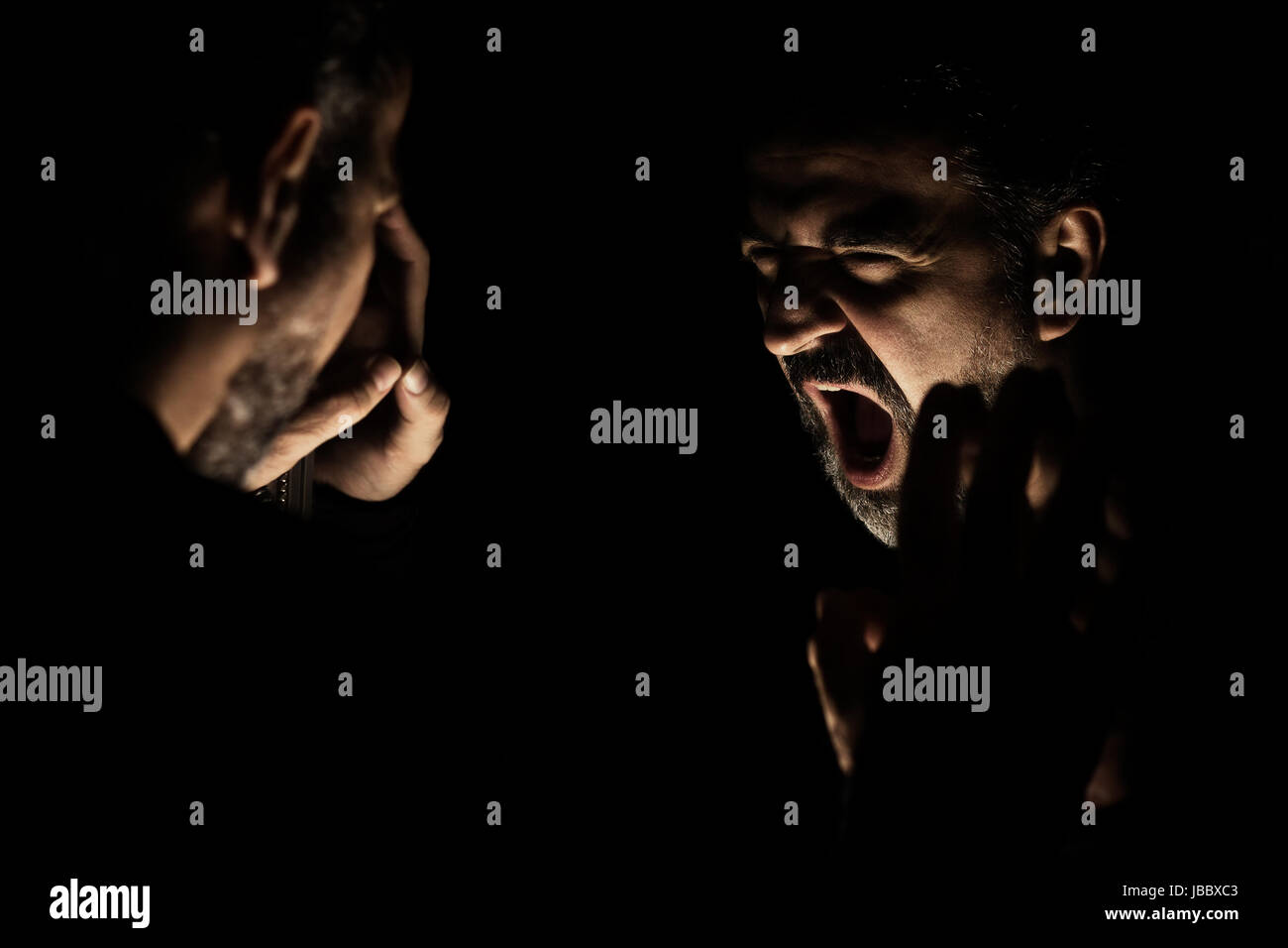 Angry man shouting in front of mirror at his reflection in a dark room ...