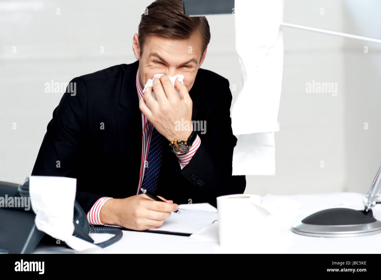 Professional sitting at desk sneezing into tissue holding pen in hand ...