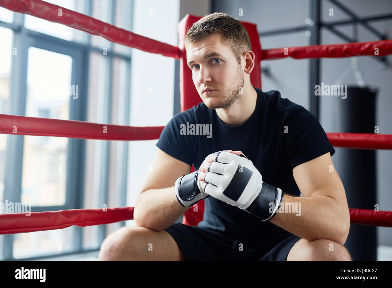 Portrait of young man posing in boxing ring sitting on floor and ...