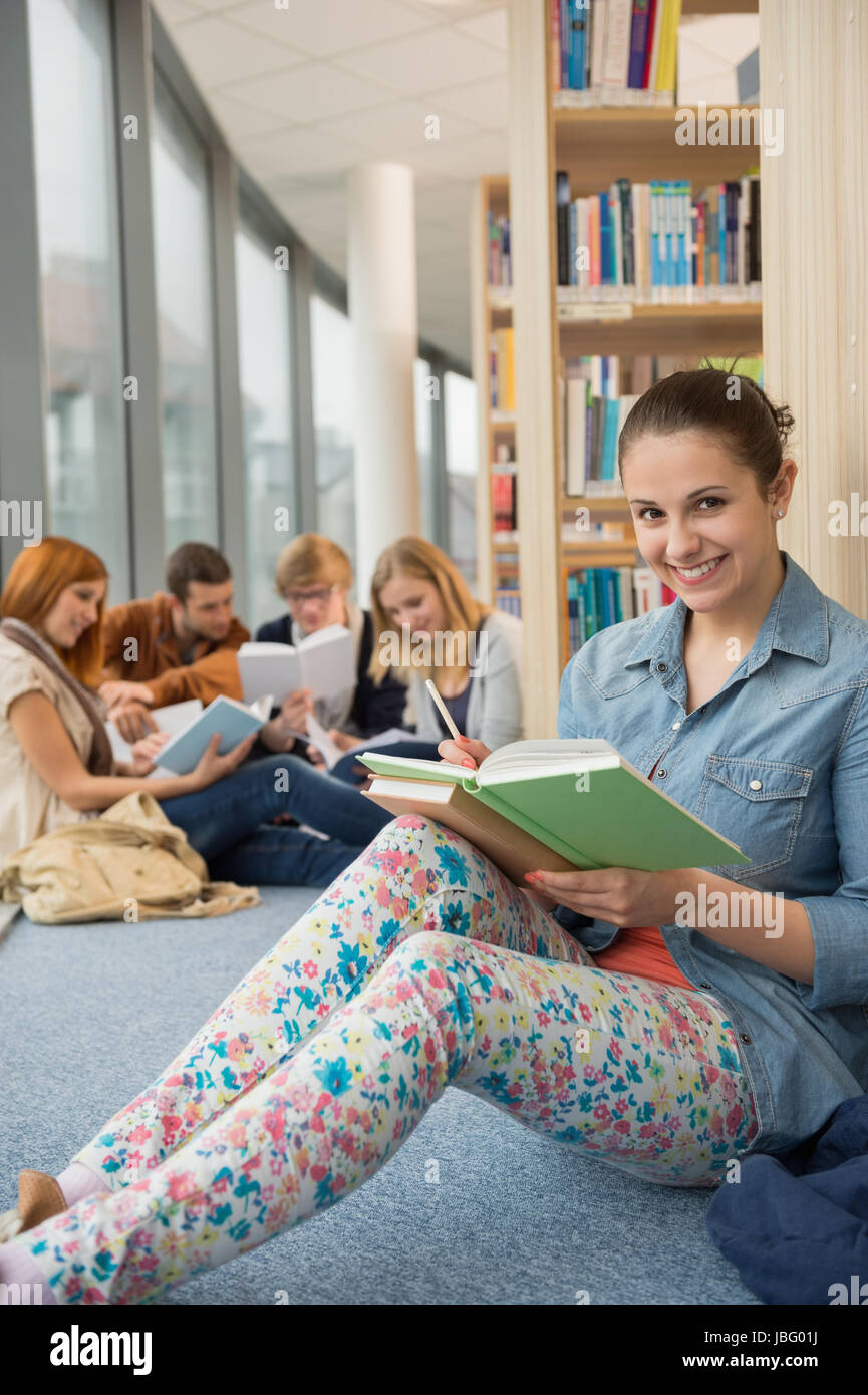 Happy student sitting in school library with classmates in background ...
