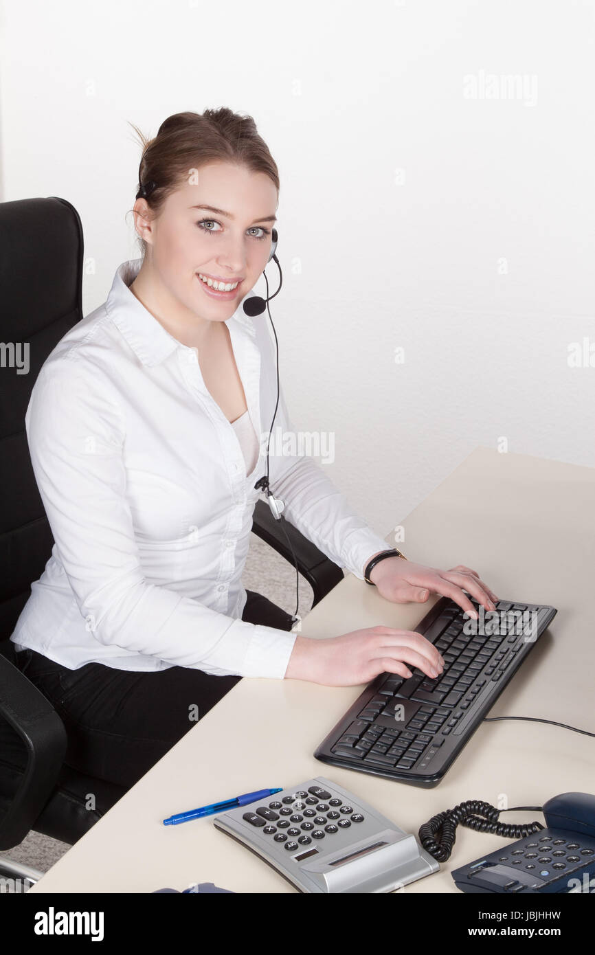Junge, lächelnde Frau mit einem Headset sitzt am Schreibtisch und tippt ...