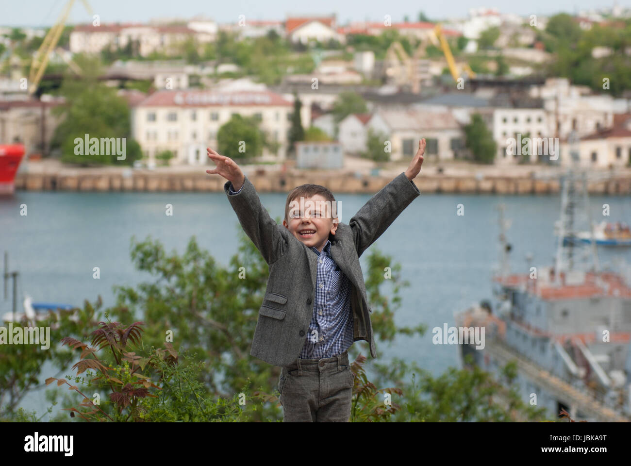 Happy handsome little boy in a jacket raised his hands up in sea port ...
