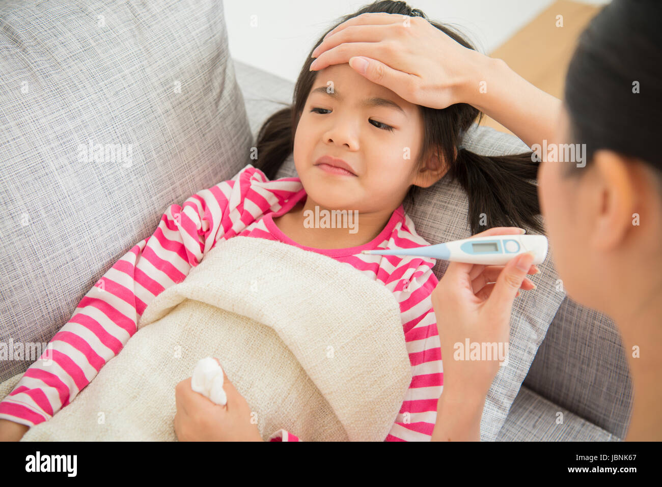 Cute little asian girl is ill lying on sofa bed mother checking a thermometer and touching ...