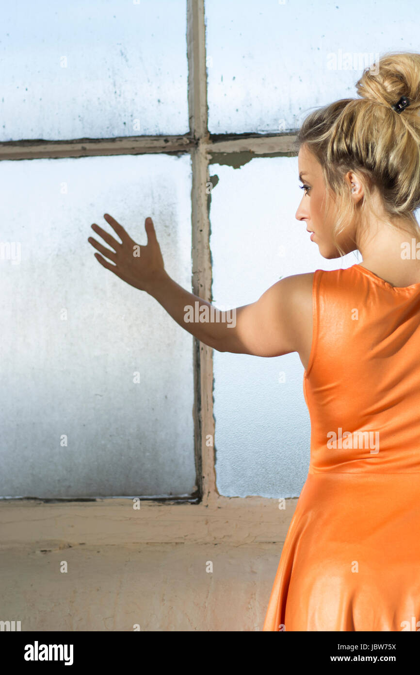 Rear view of a young blond woman standing by the window hand touching ...