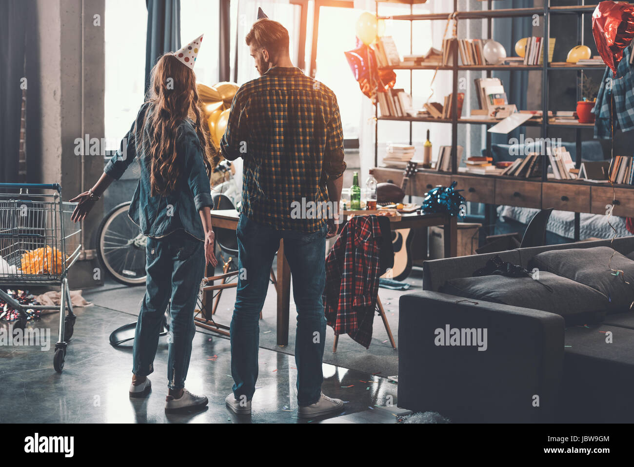 back view of couple in birthday hats standing in messy room after party ...