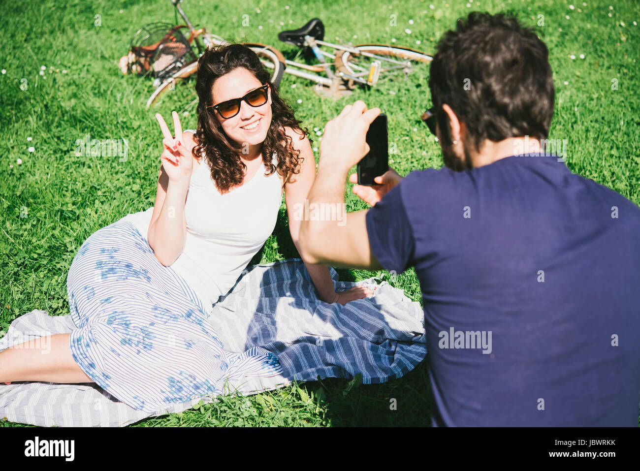 Over shoulder view of man photographing girlfriend sitting in park ...