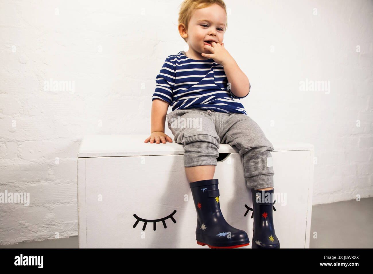 Portrait of male toddler sitting on toy chest with fingers in mouth ...