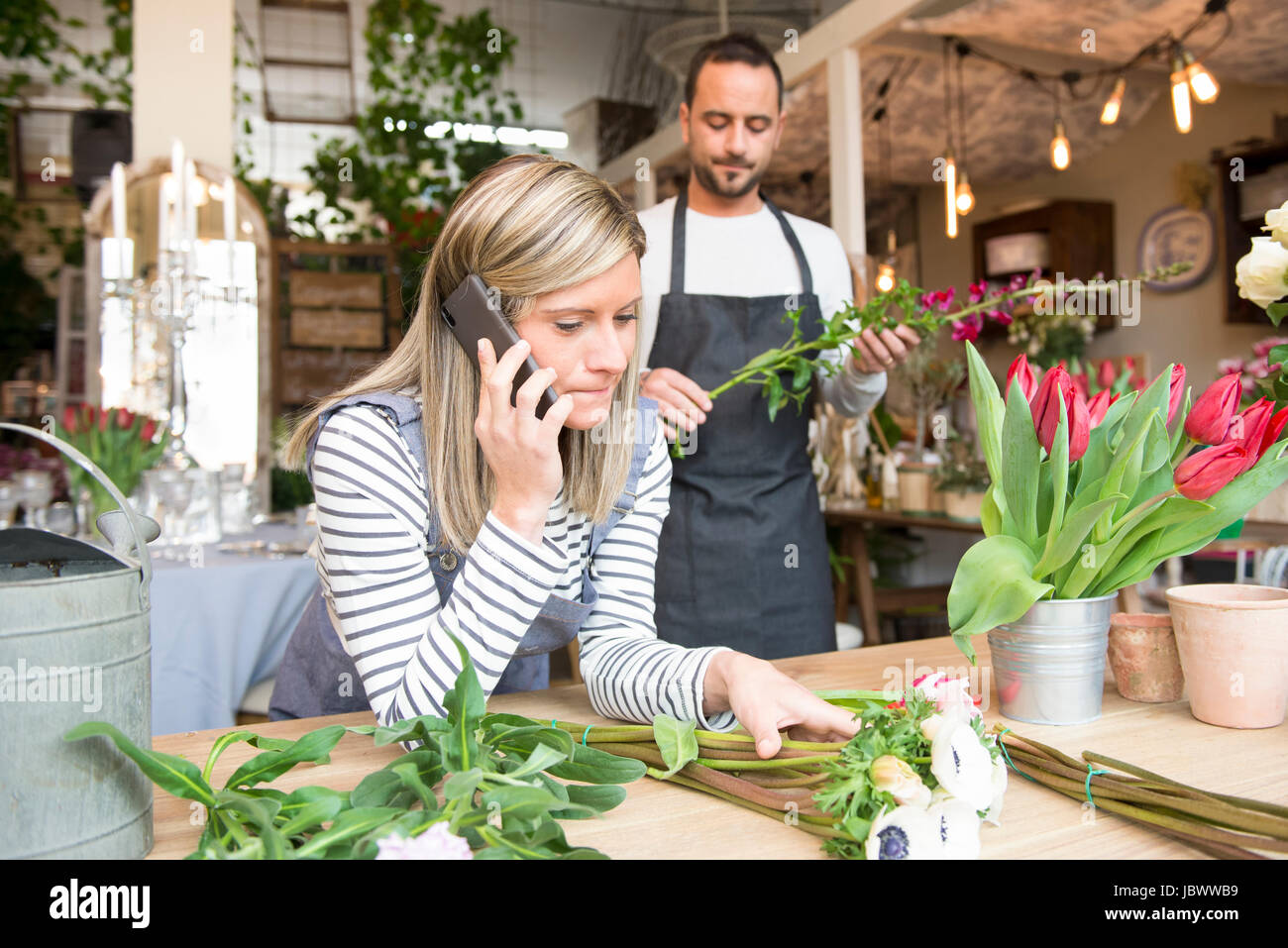 Female florist talking on smartphone, male colleague preparing flower order behind her Stock ...