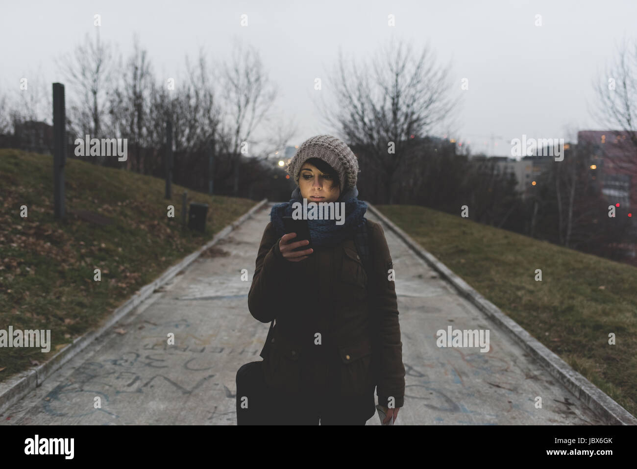 Female backpacker walking in city park at dusk looking at smartphone ...