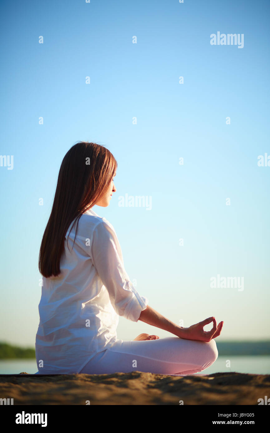 Side view of meditating woman sitting in pose of lotus against blue sky ...