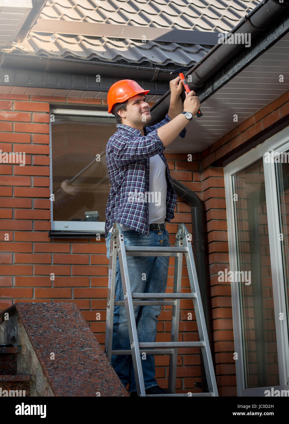 Young worker standing on step ladder and repairing gutter on house ...