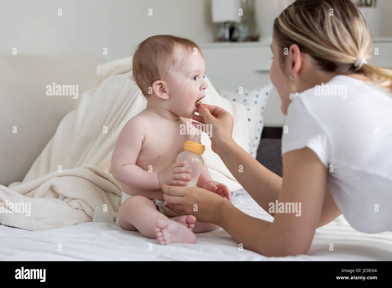 Young mother giving baby cookies to her 9 months old son sitting on bed ...