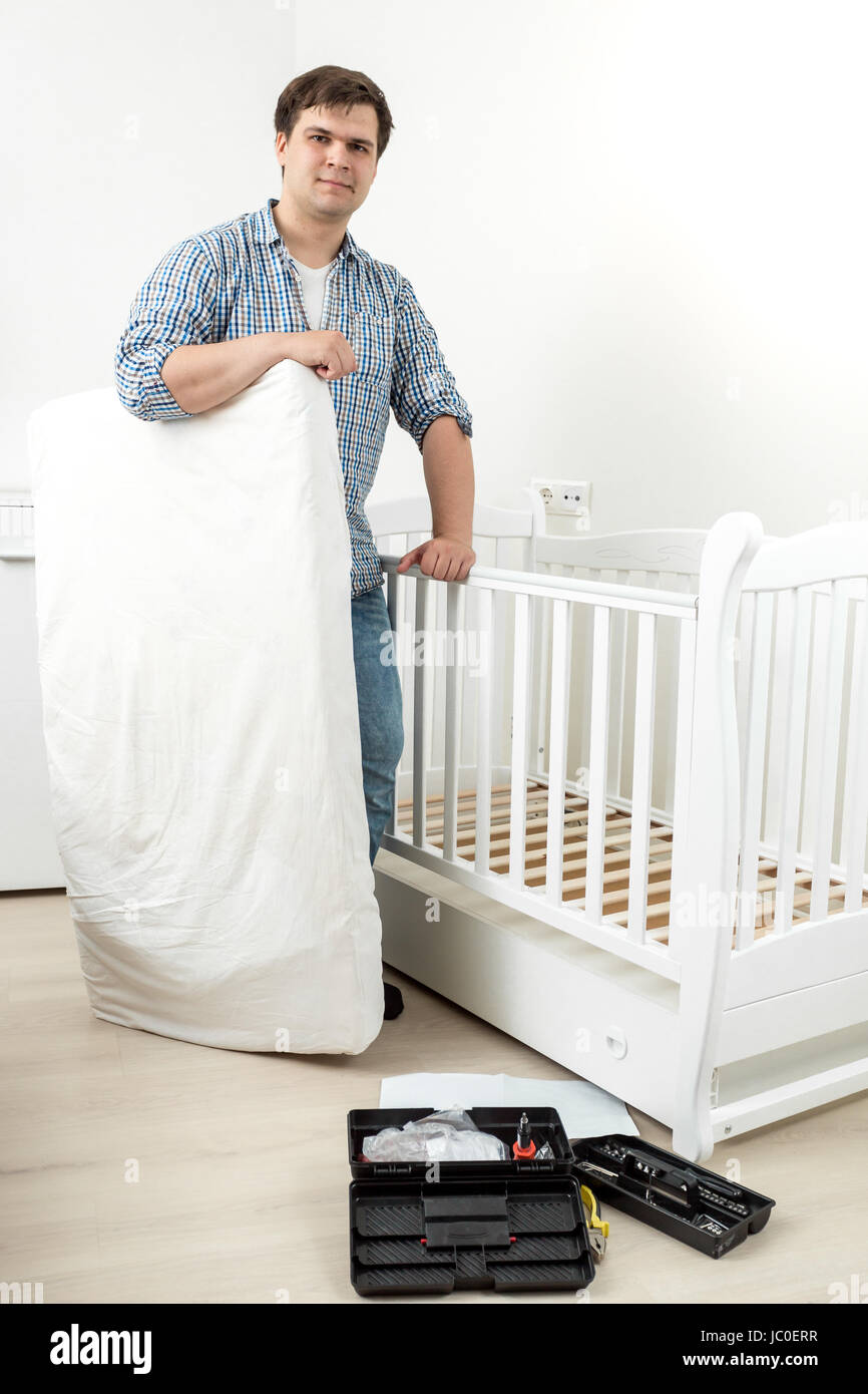 Young smiling man standing with mattress at disassembled baby's cot ...