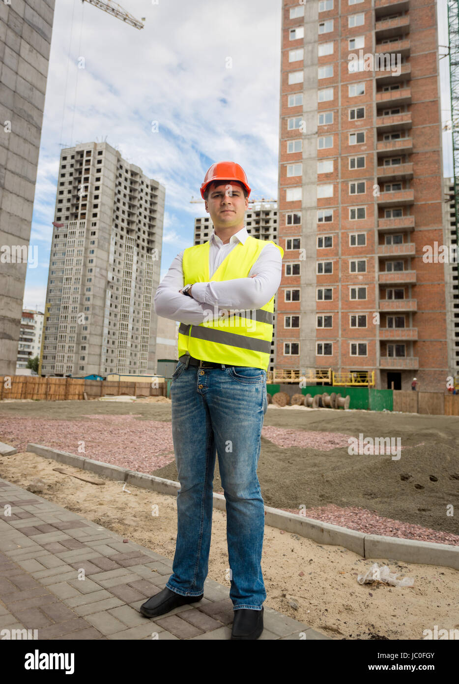 Smiling confident worker in helmet and safety vest standing on building ...