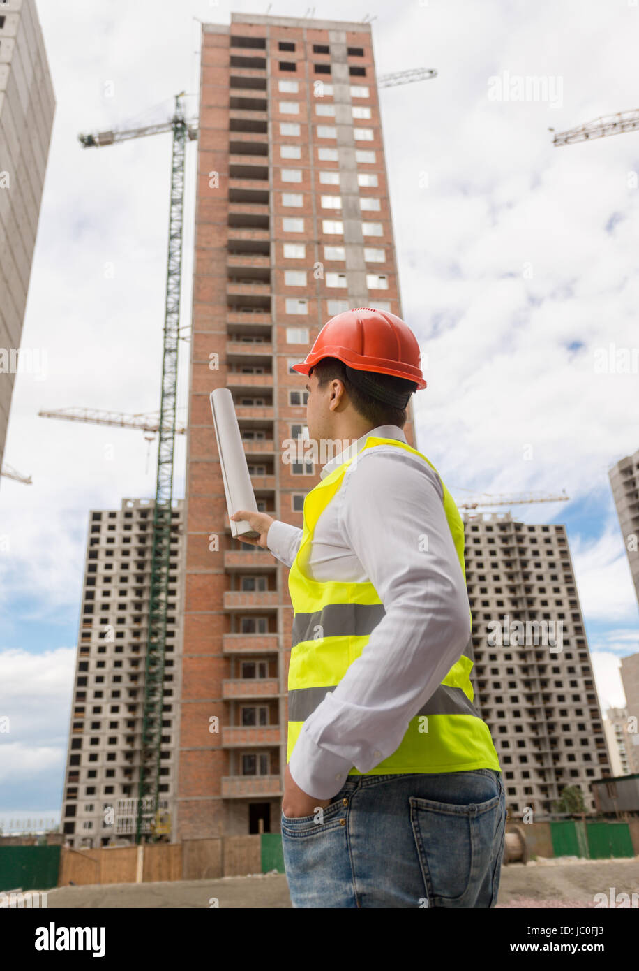 Rear view of young architect pointing with blueprints at building under ...
