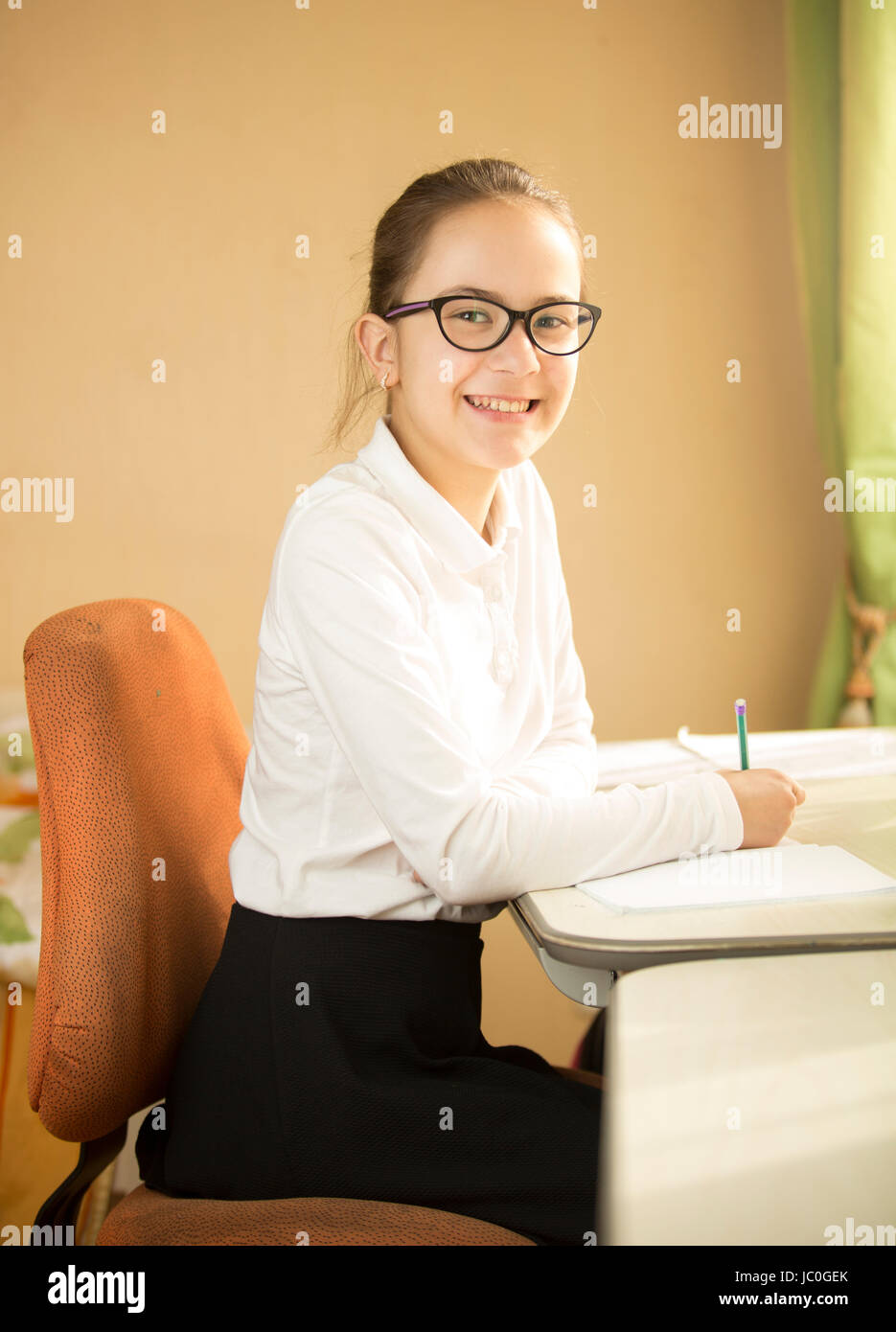 Portrait of beautiful schoolgirl wearing eyeglasses posing behind desk ...