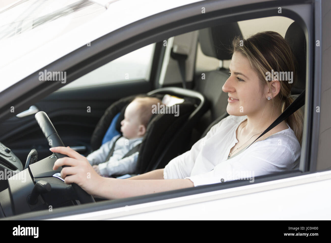 Portrait of young mother driving car with her little baby on front seat ...