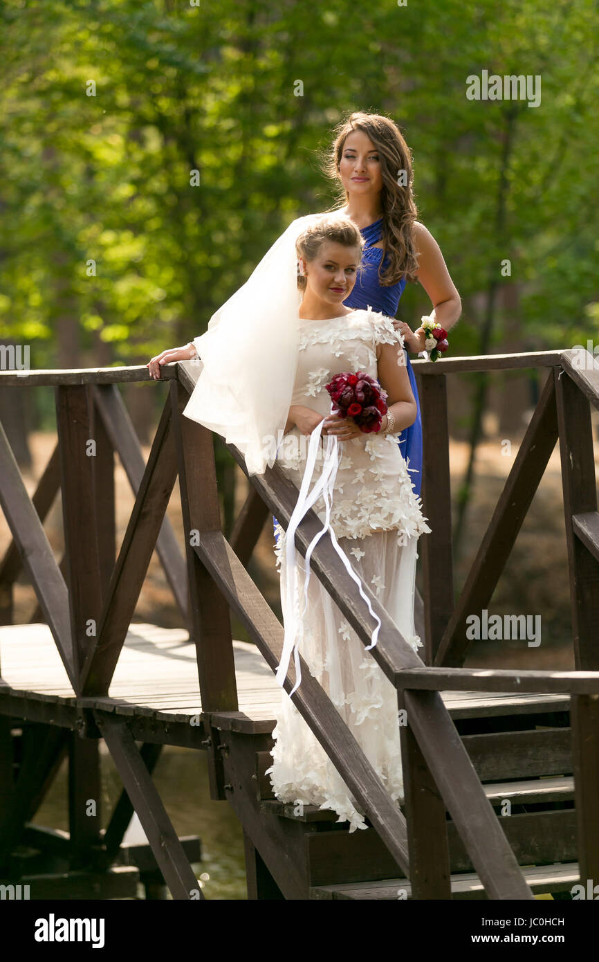Beautiful bride hugging with bridesmaid on wooden bridge at windy day ...