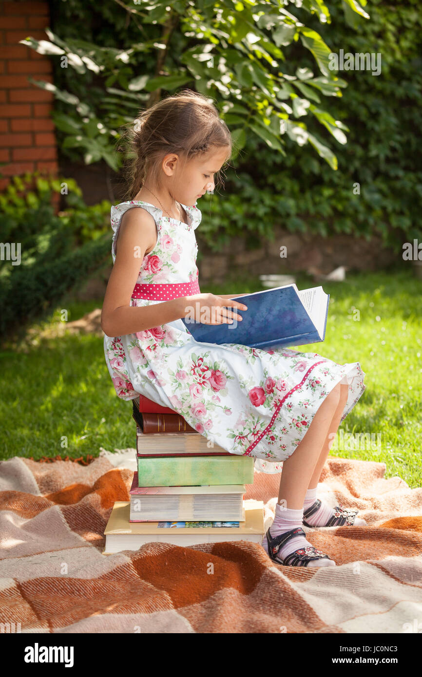 Little girl in cute dress sitting on pile of books and reading Stock ...