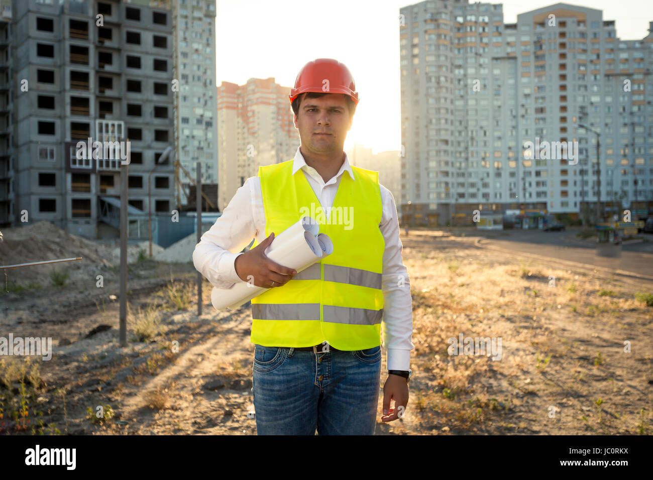 Portrait of young engineer posing with blueprints against building site ...