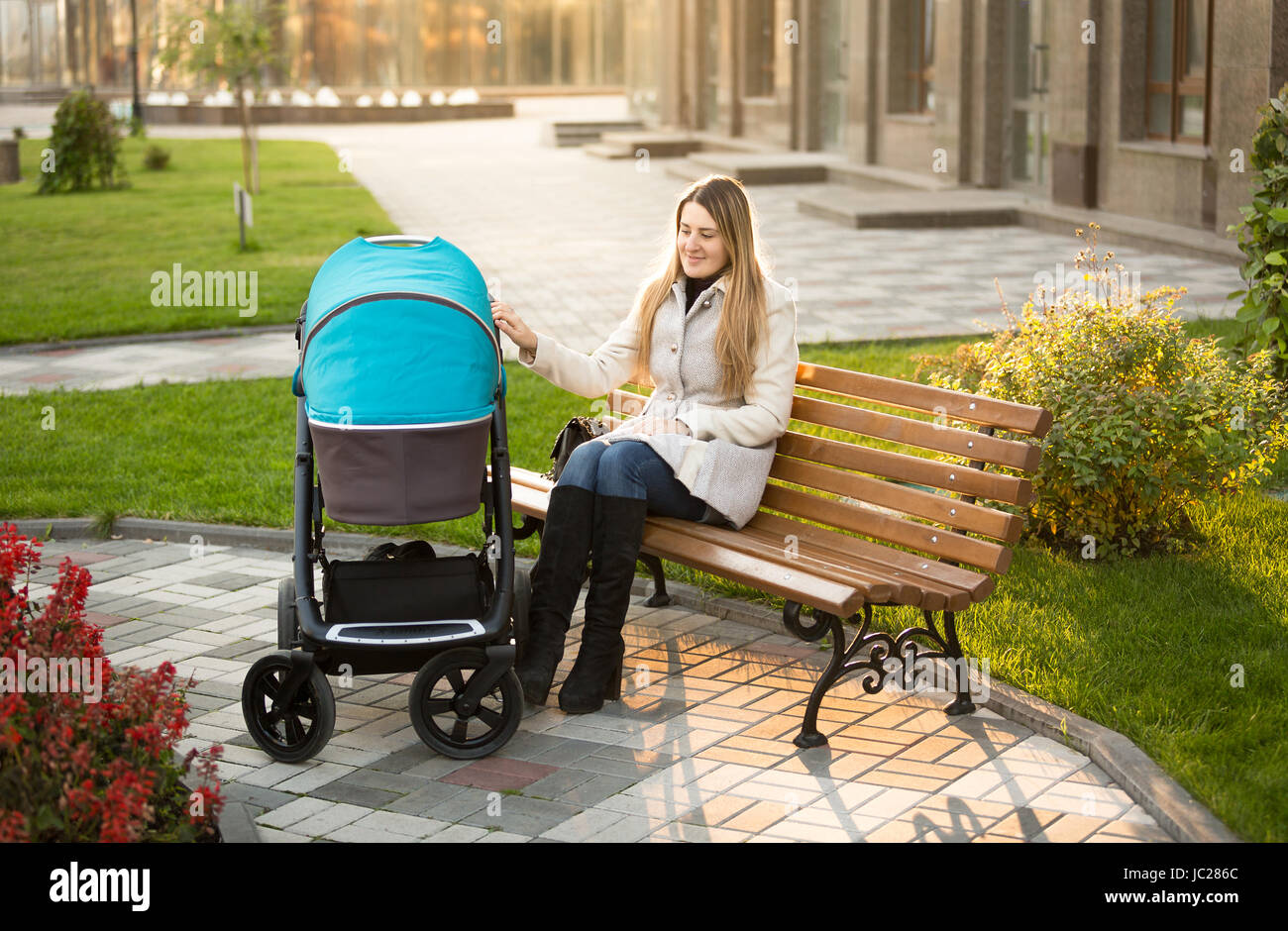 Beautiful mother sitting on bench at park and swaying baby stroller ...