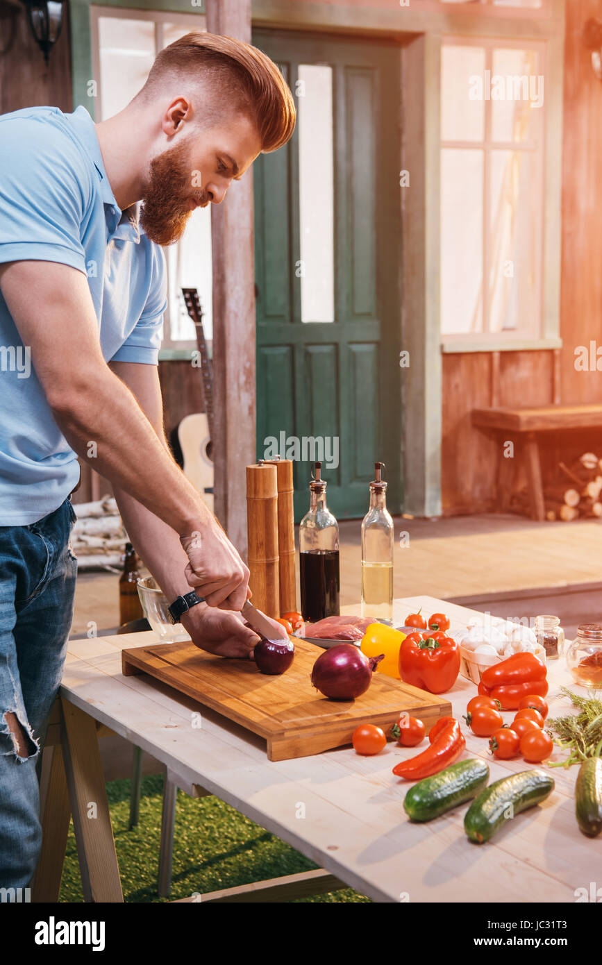 Side view of bearded young man cutting onion for barbecue Stock Photo ...