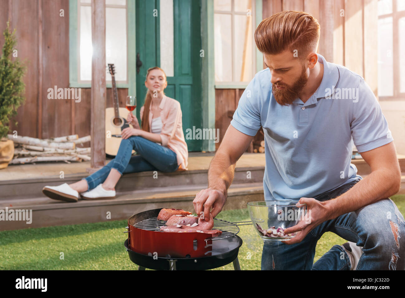 bearded man roasting meat on barbecue grill with woman with wine behind ...
