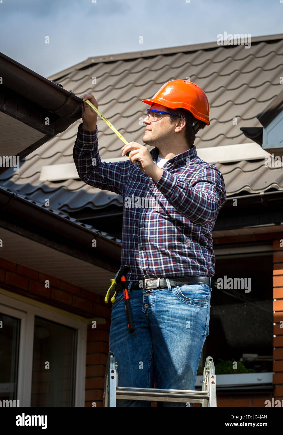 Young handyman standing on high ladder and measuring roof with tape ...