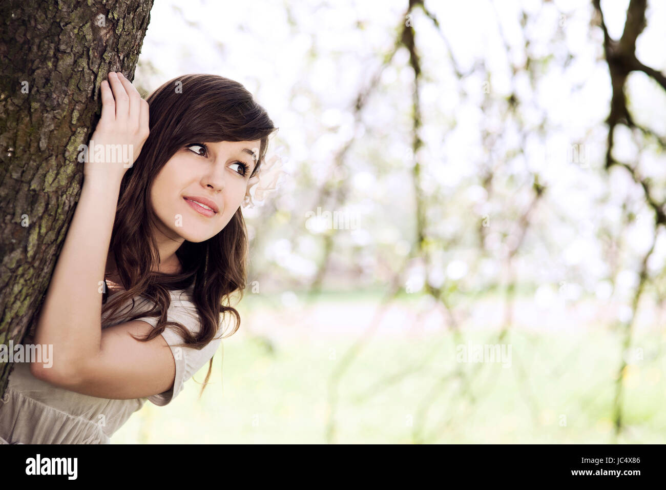Beautiful young woman hiding behind tree trunk Stock Photo - Alamy