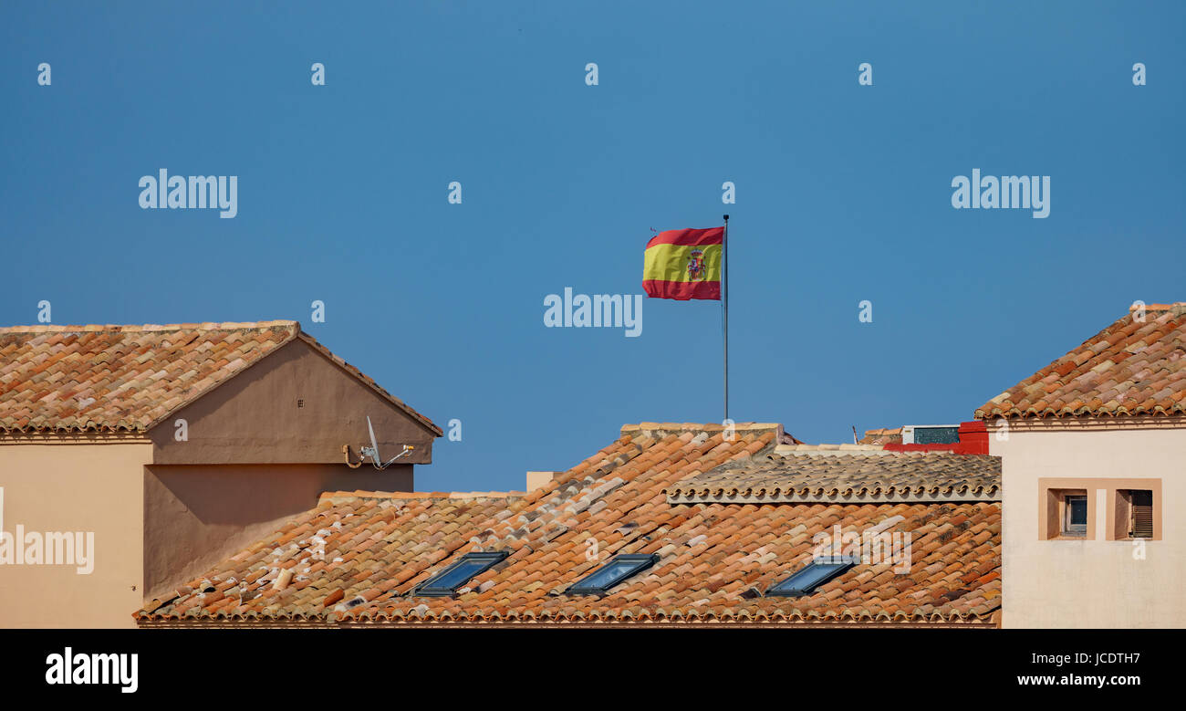 Long shot of traditional tile roofs with spanish flag on top over blue ...